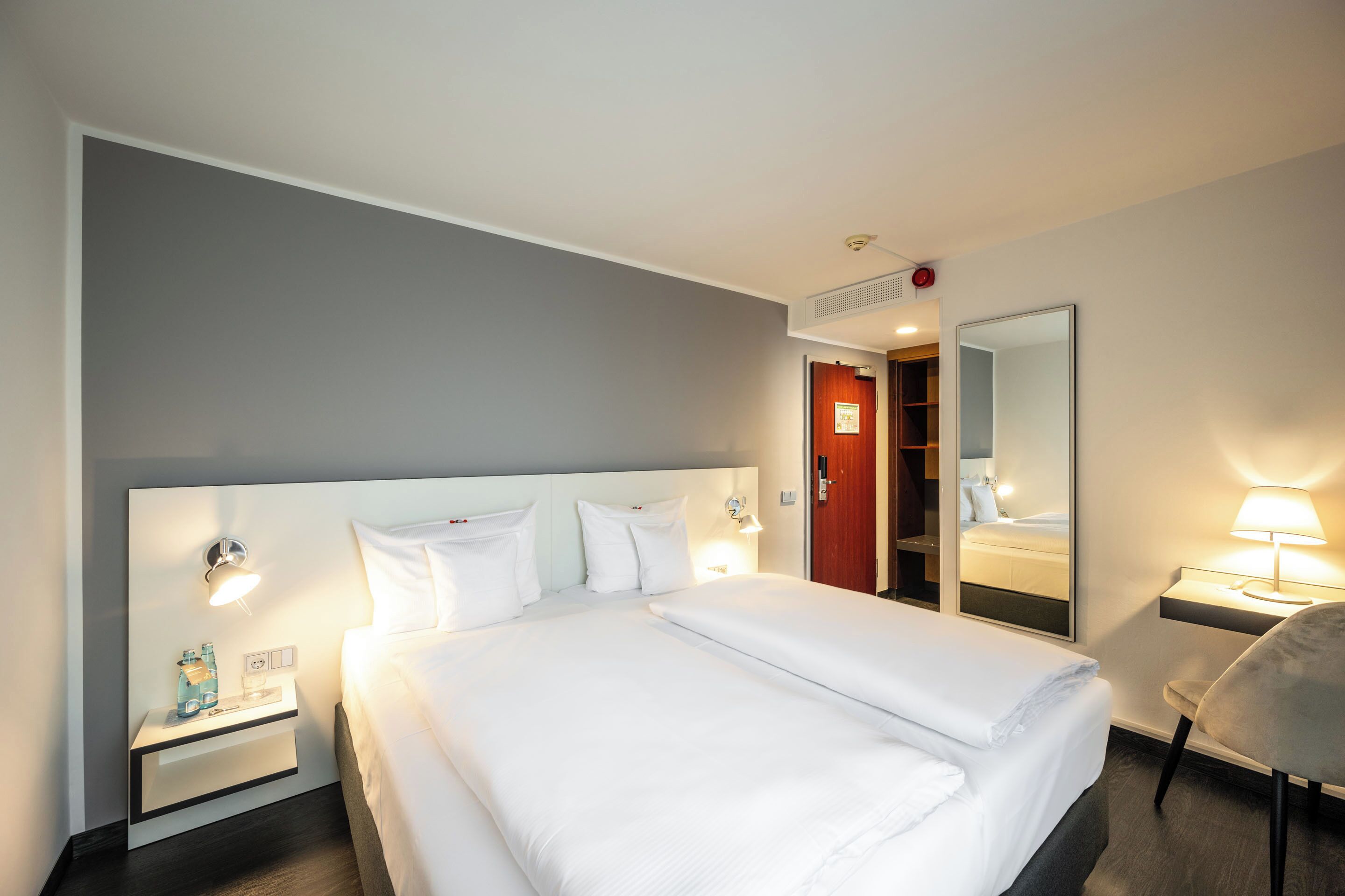 Quarto de hotel com uma cama grande, mesas de cabeceira, um espelho e uma escrivaninha com abajur.