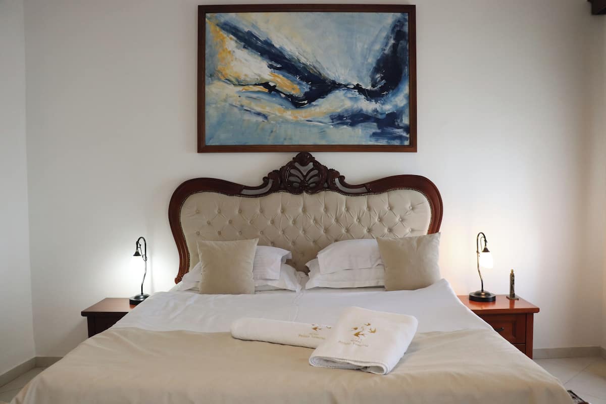 Uma cama bem arrumada com edredom branco, duas luminárias de mesa e um quadro abstrato emoldurado acima.