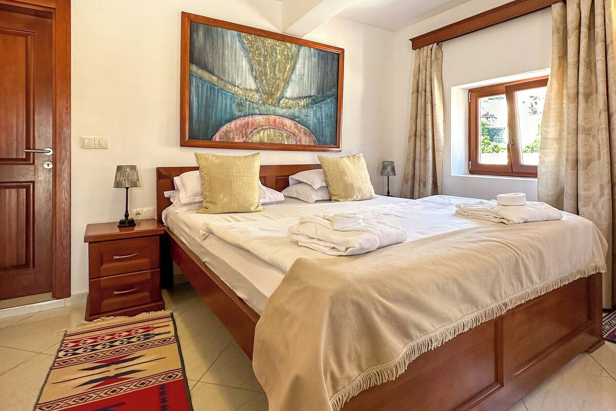 Quarto com uma cama grande, mesa de cabeceira de madeira, luminária, quadro na parede e janela com cortinas.