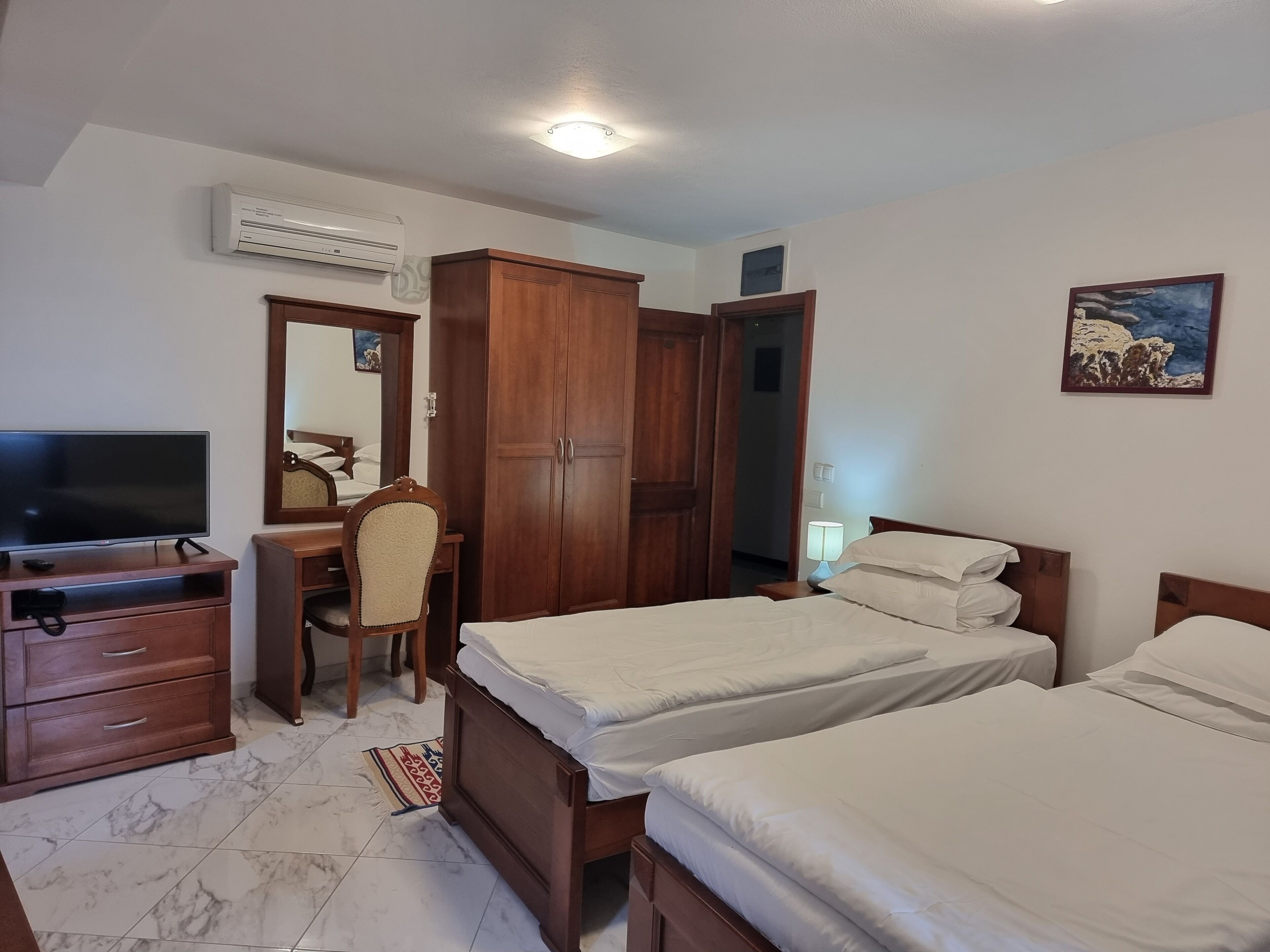 Quarto de hotel com duas camas, uma escrivaninha com cadeira, televisão, guarda-roupa e ar condicionado.