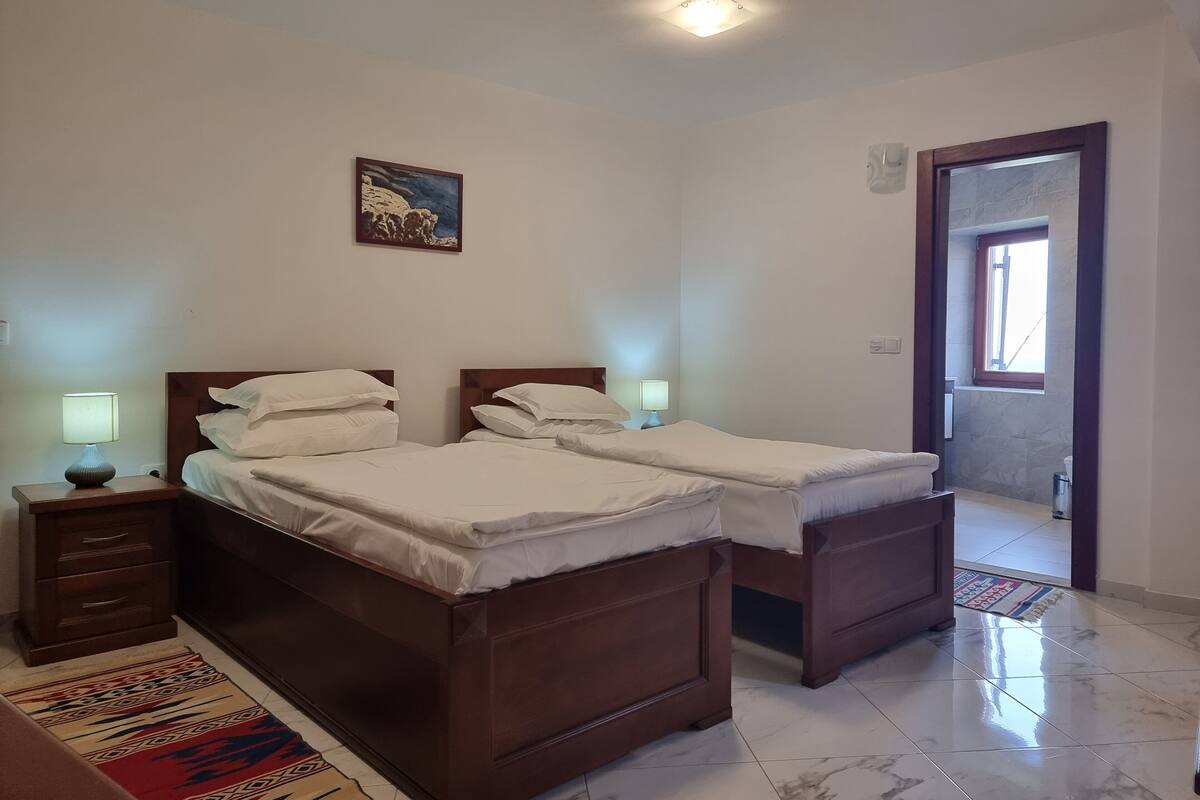 Quarto com duas camas, mesa de cabeceira com abajur, quadro na parede e janela com vista.