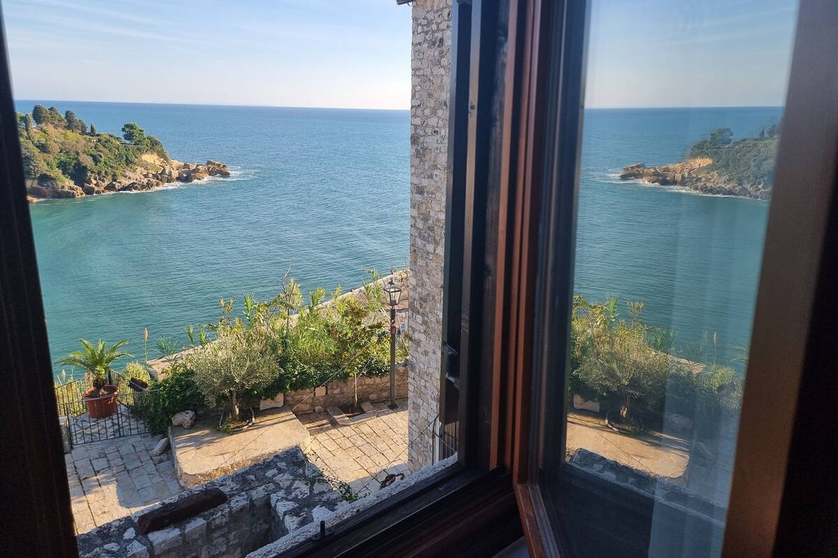 Vista costeira numa janela com escadaria de pedra que dá para um terraço com plantas em vasos e vista para o mar.