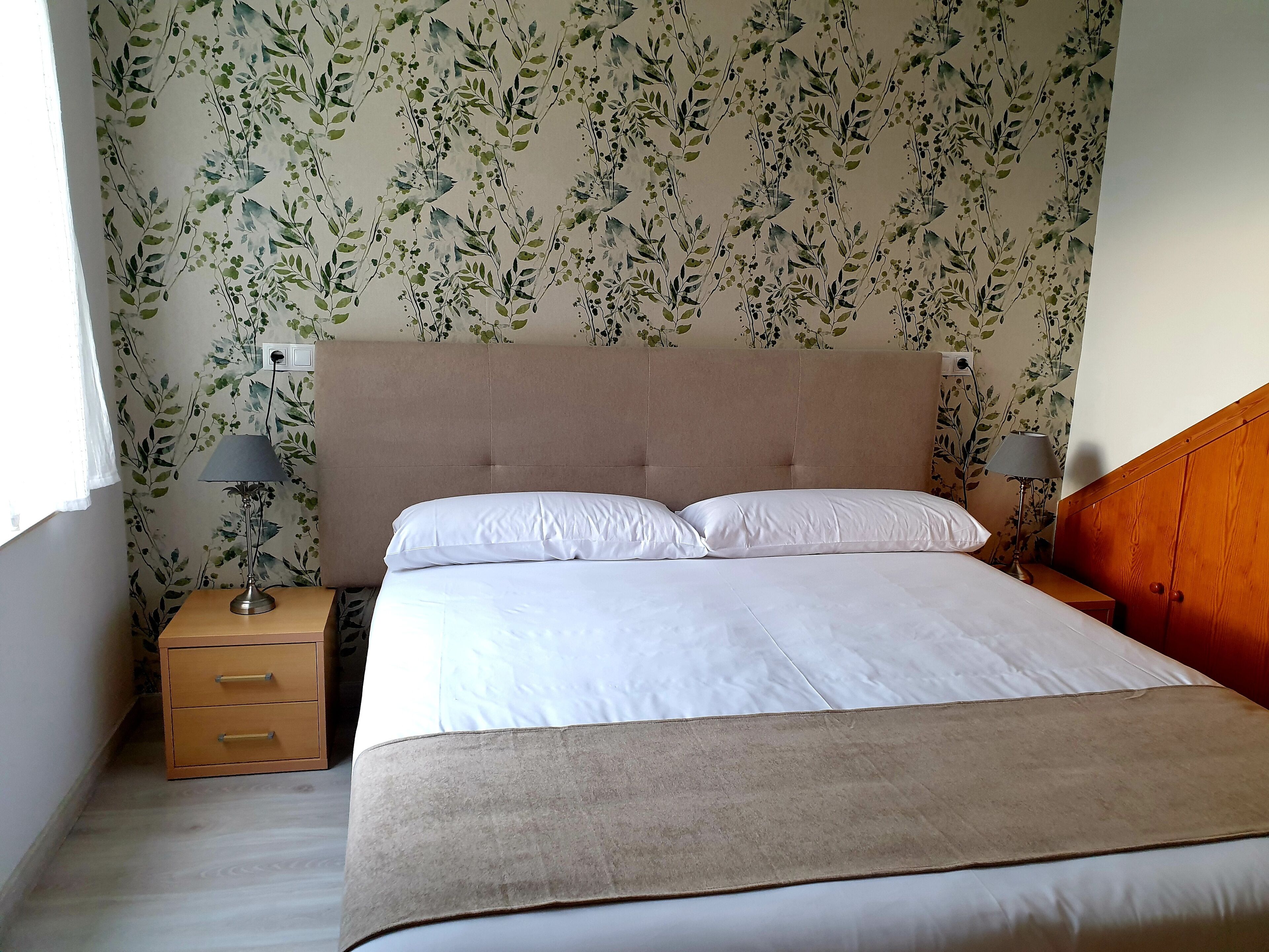 Cama bem arrumada com lençóis brancos, mesa de cabeceira de madeira com abajur e parede com papel de parede estampado com motivos florais.