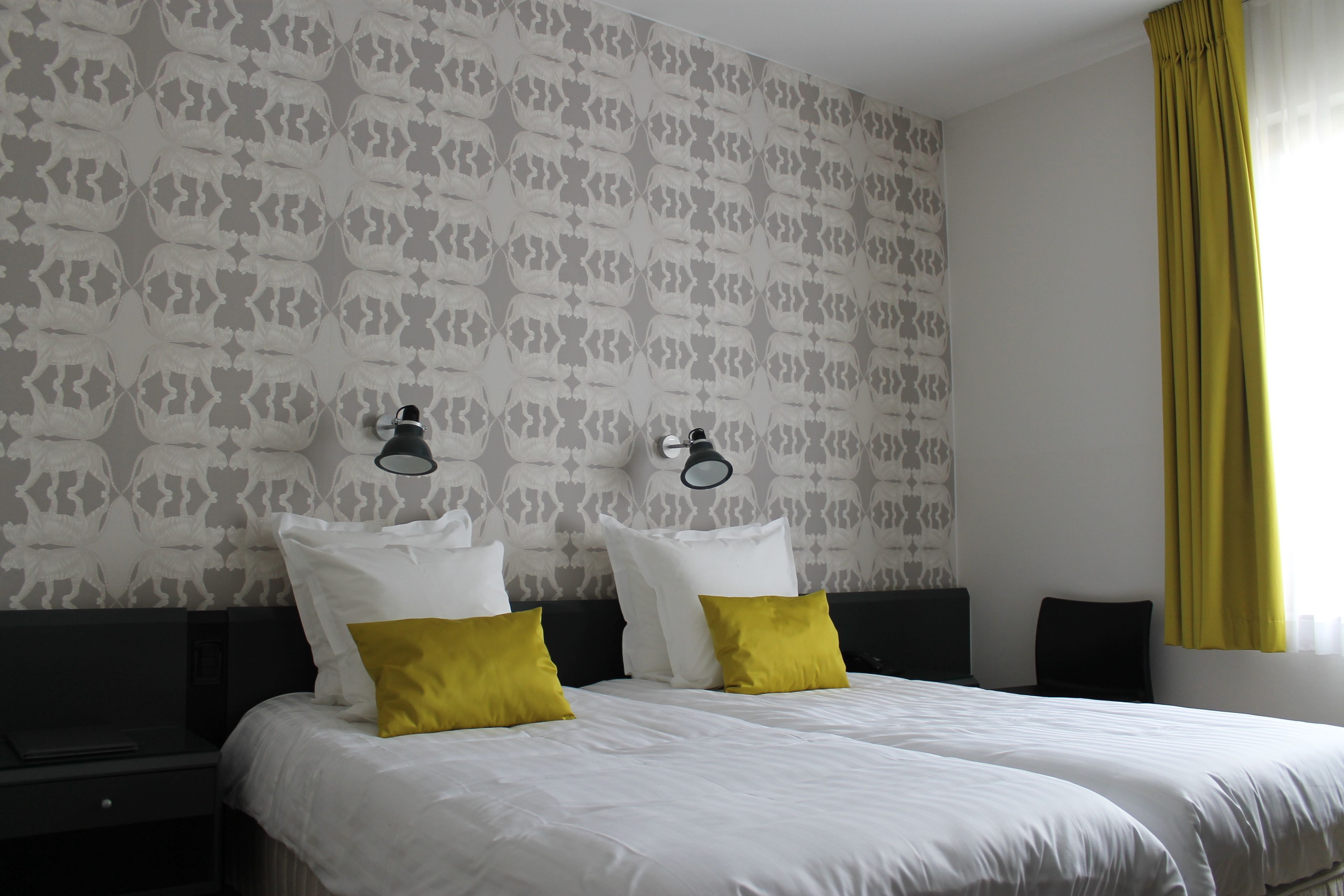 Quarto de hotel com papel de parede estampado, uma cama com almofadas brancas e amarelas, uma mesa de cabeceira e uma janela com cortinas amarelas.