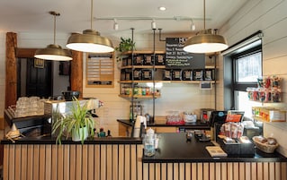 Interior de uma cafeteria com um quadro de cardápio, máquinas de café e diversos alimentos nas prateleiras.
