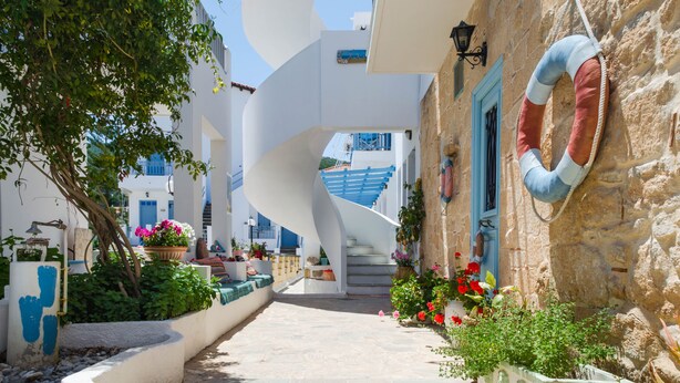 Um caminho estreito ladeado por edifícios brancos com detalhes azuis, conduzindo a uma escadaria em espiral.