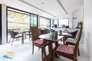 Uma sala de jantar espaçosa com mesas e cadeiras de madeira, grandes janelas com molduras verdes e vista para o verde lá fora.