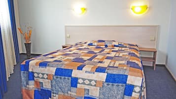 Cama bem arrumada com colcha estampada, duas luminárias de cabeceira e uma mesinha ao lado da cama.