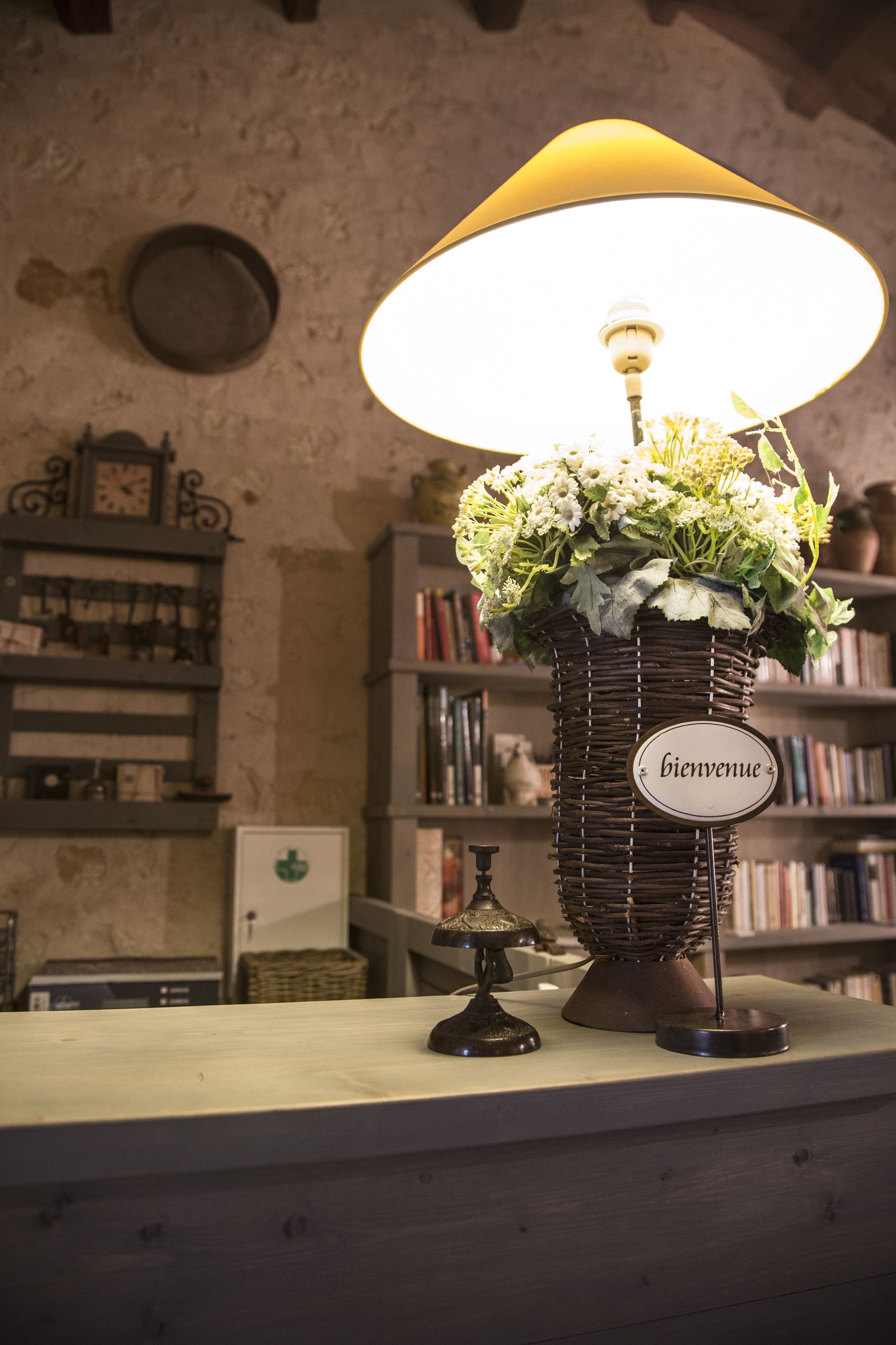 Uma luminária com a placa 'bienvenue', um cesto de flores e uma estante de livros ao fundo.