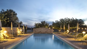 Piscina retangular com parede de pedra ao fundo, rodeada por espreguiçadeiras e guarda-sóis.