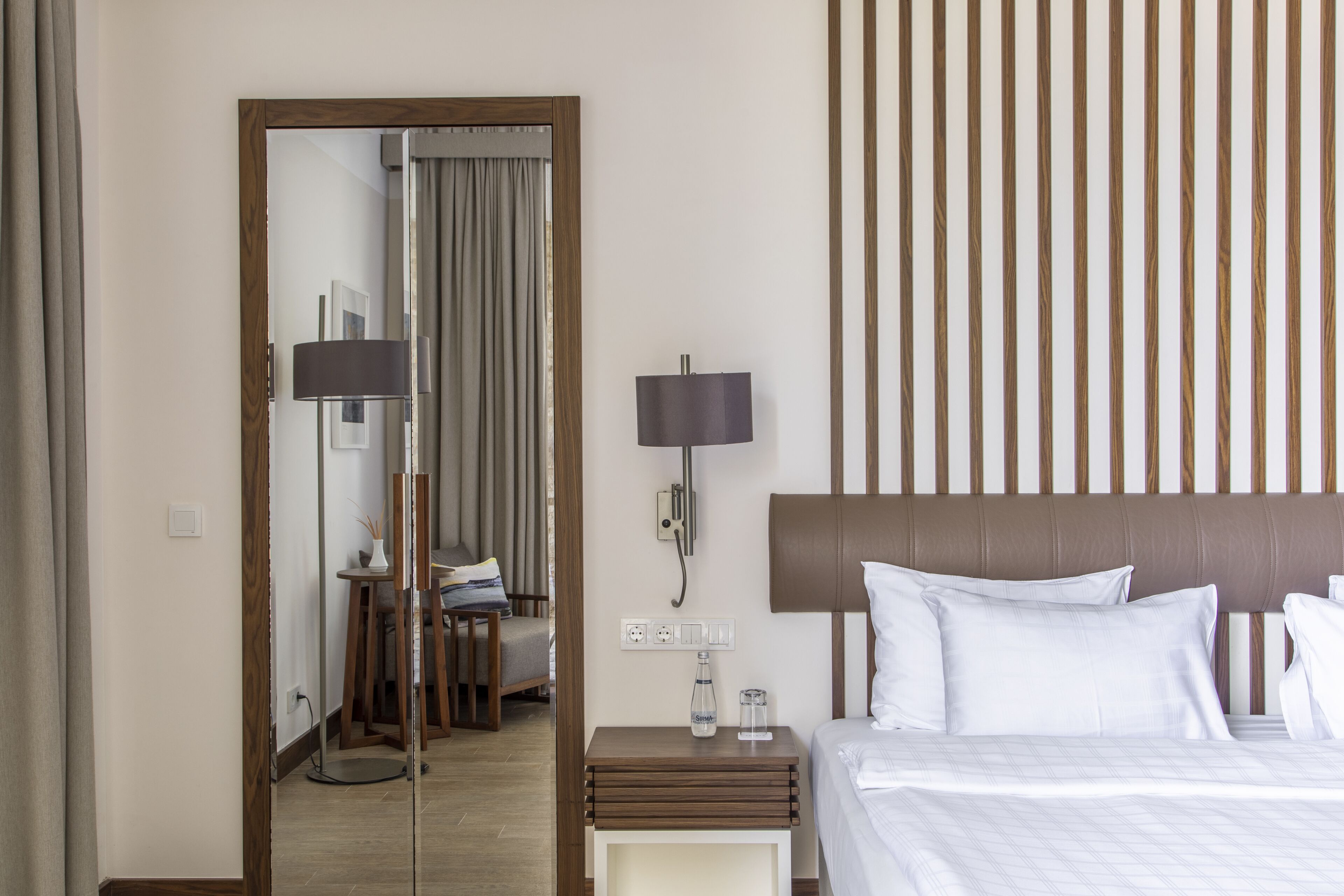 Quarto de hotel com cama, luminárias, mesa de cabeceira, espelho e vista para o corredor.