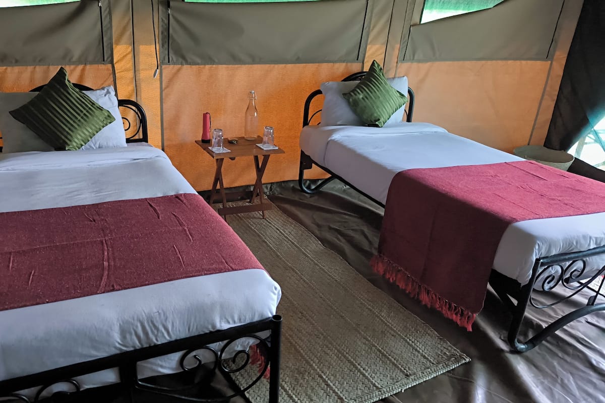 Quarto em alojamento tipo tenda com duas camas, cada uma com colcha branca e vermelha, almofadas verdes e uma pequena mesa com uma garrafa e copos.