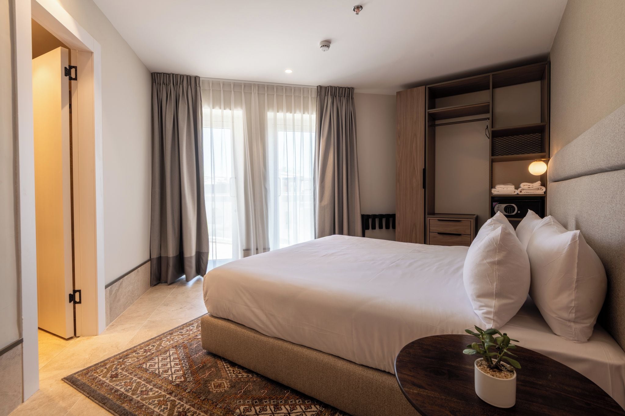 Quarto de hotel com uma cama grande, mesa de cabeceira com uma planta, guarda-roupa e mesa de apoio com abajur.