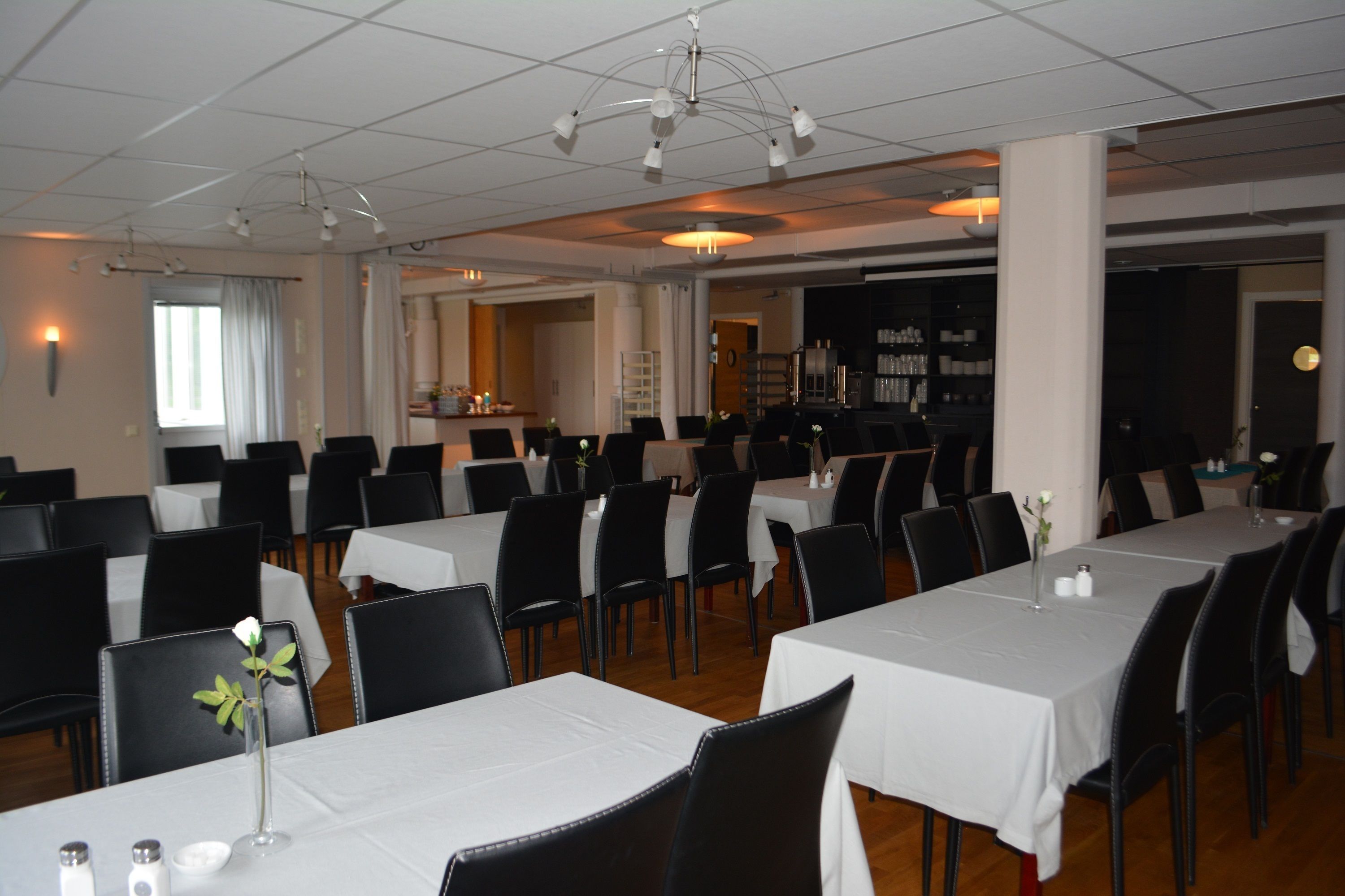 Uma sala de jantar com várias mesas e cadeiras, um candelabro e uma cozinha ao fundo.