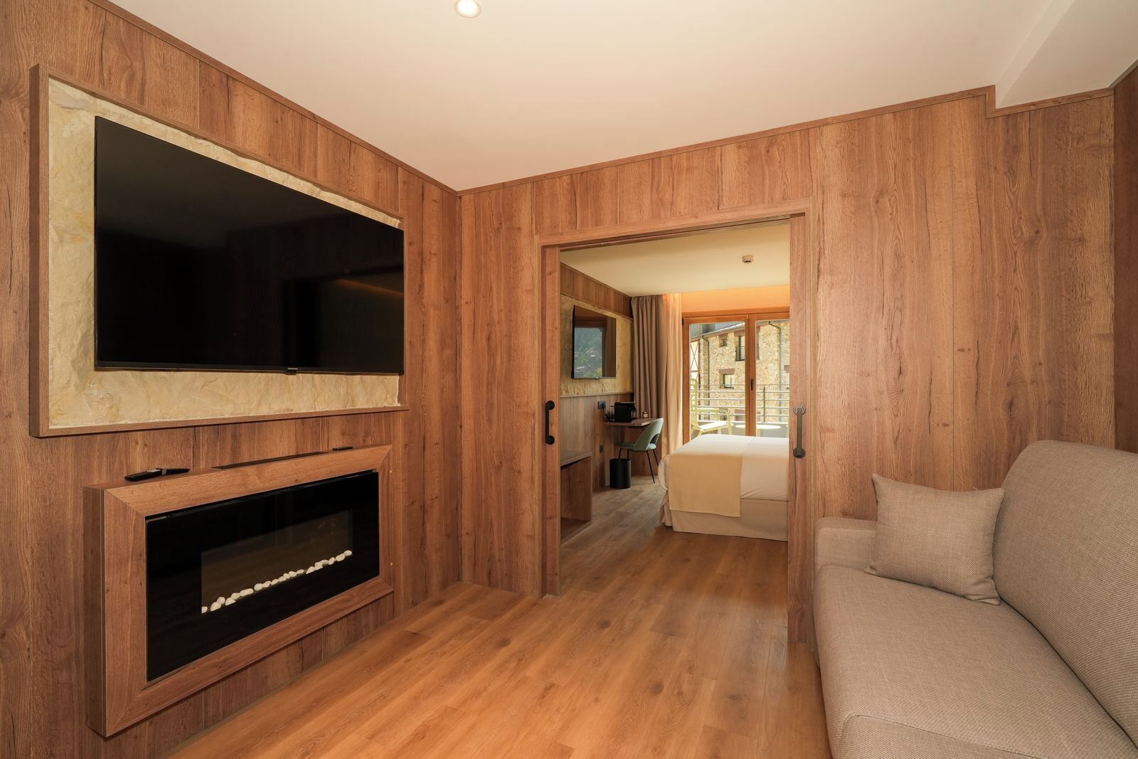 Quarto de hotel com piso de madeira, televisão de tela plana, lareira, sofá e varanda com mesa e cadeiras.