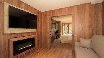Quarto de hotel com piso de madeira, televisão de tela plana, lareira, sofá e varanda com mesa e cadeiras.