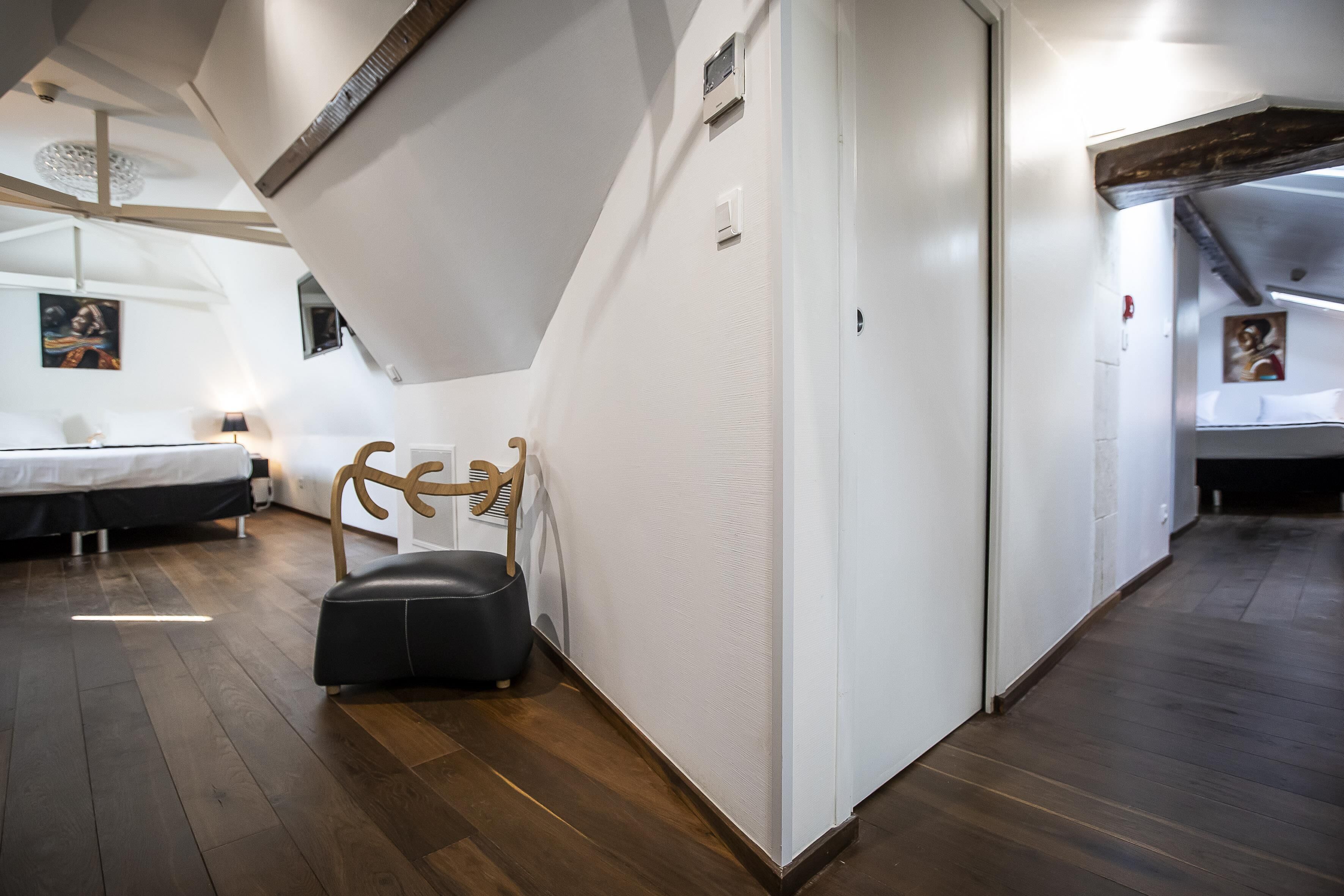 Quarto de hotel moderno com piso de madeira, uma poltrona de couro preto, uma cama e uma parede branca com um quadro.