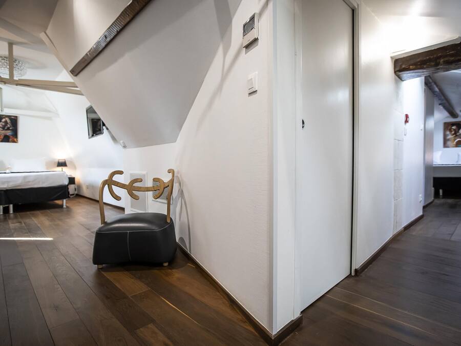 Quarto de hotel moderno com piso de madeira, uma poltrona de couro preto, uma cama e uma parede branca com um quadro.