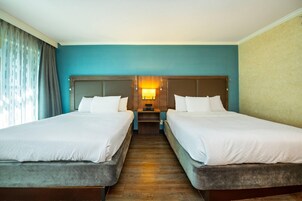 Quarto de hotel com duas camas, cabeceira de madeira, mesa de cabeceira com abajur e janela com cortinas.