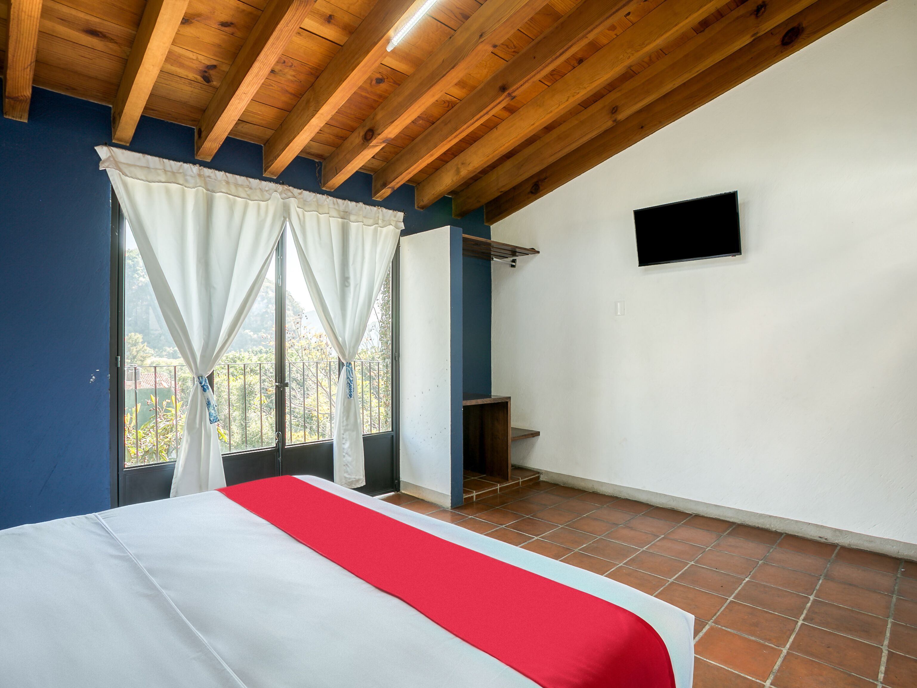 Quarto com cama, janela com cortinas, televisão na parede e varanda com guarda-corrimão.