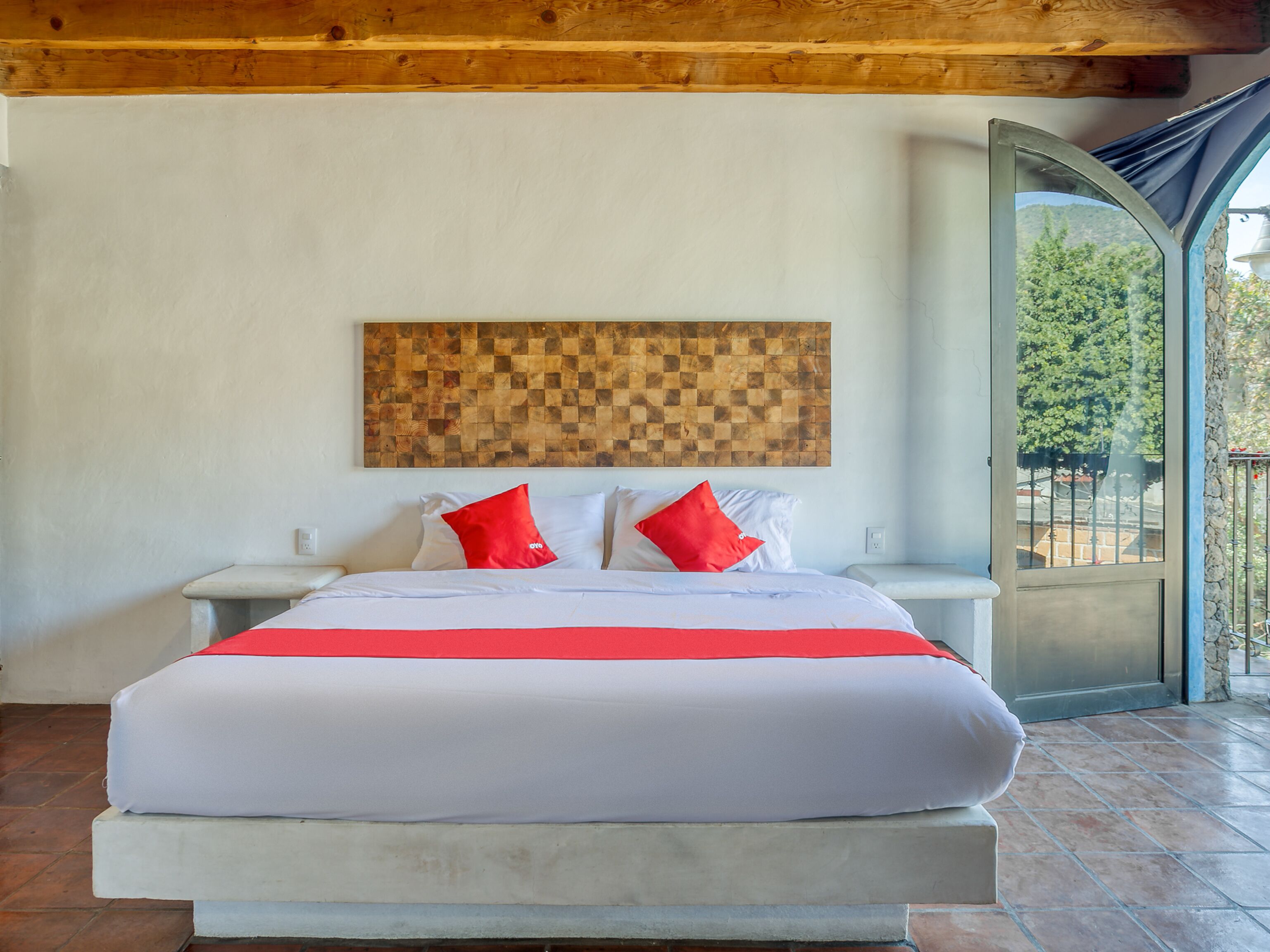 Quarto com uma cama grande, roupa de cama branca e almofadas vermelhas. Possui cabeceira de madeira, parede de pedra e vista para o verde através de uma porta de vidro.
