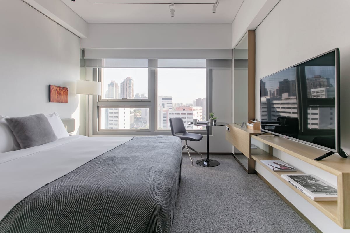 Quarto de hotel com uma cama, escrivaninha, cadeira, televisão e uma grande janela com vista para a cidade.