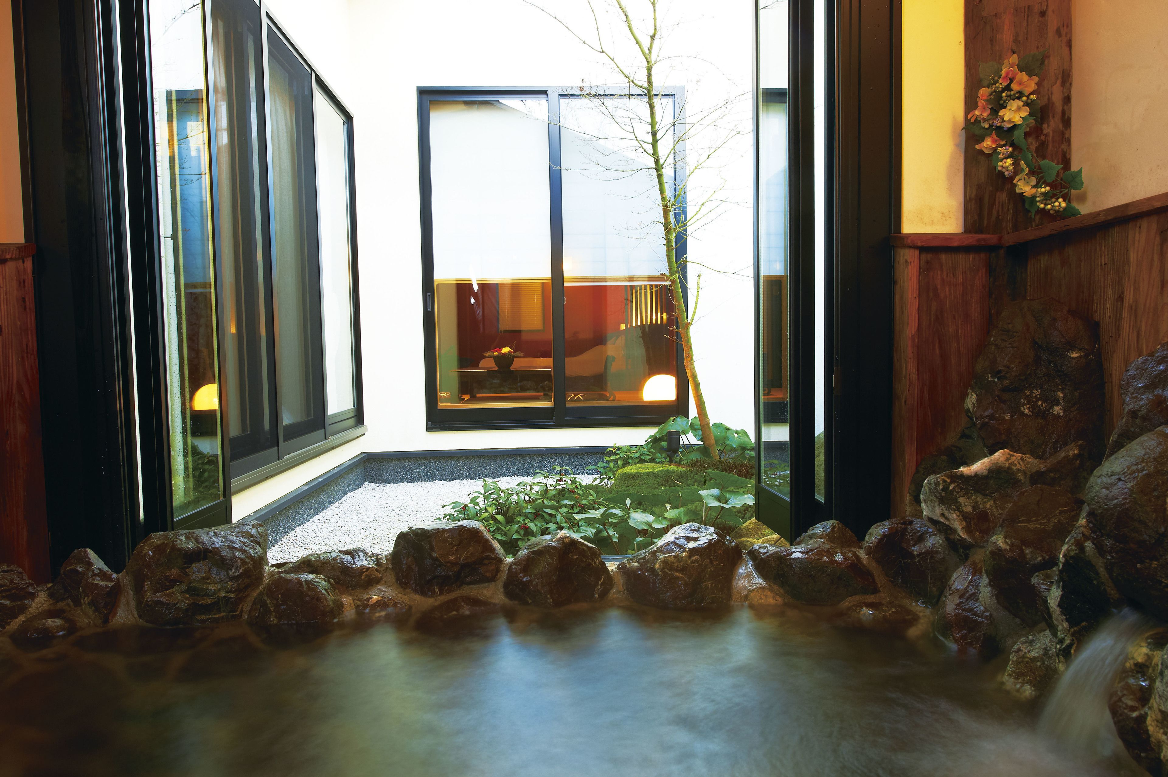 Interior moderno com uma fonte de pedra, um banco de madeira e vista através de portas de vidro para um quarto com cama e planta.