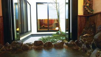 Interior moderno com uma fonte de pedra, um banco de madeira e vista através de portas de vidro para um quarto com cama e planta.