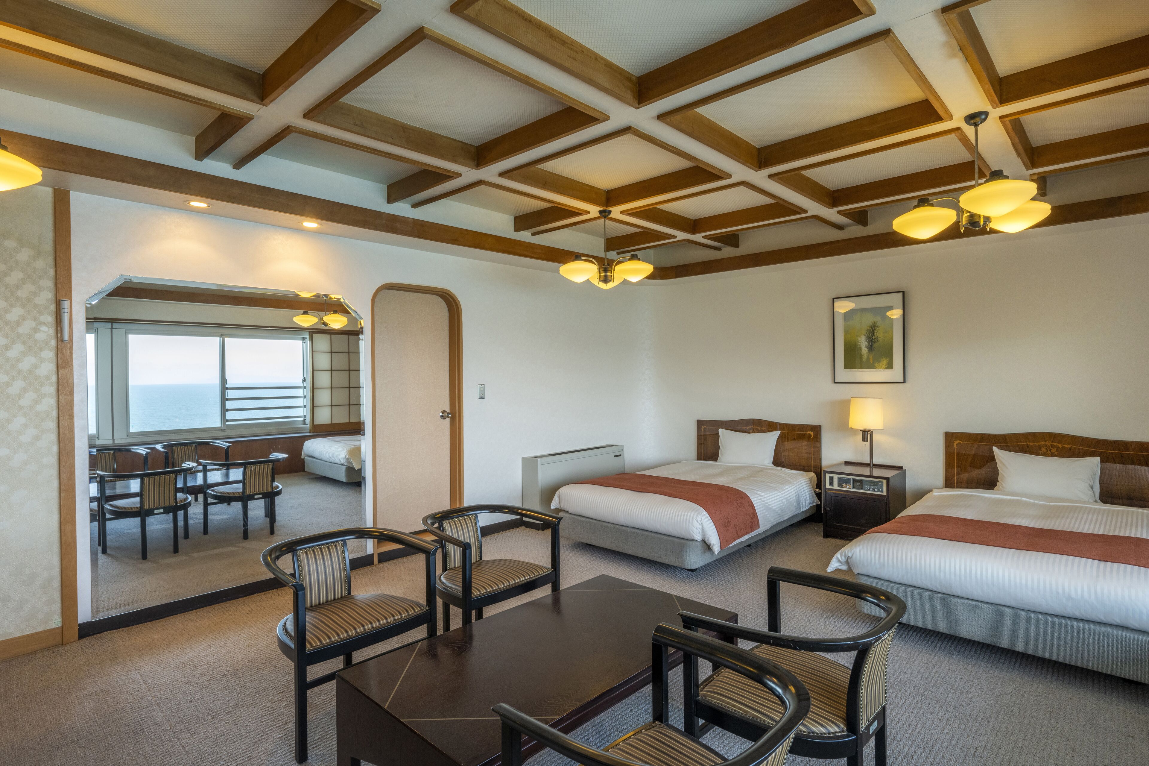 Quarto de hotel com duas camas, área de refeições com cadeiras, uma mesa e vista para o mar através de grandes janelas.