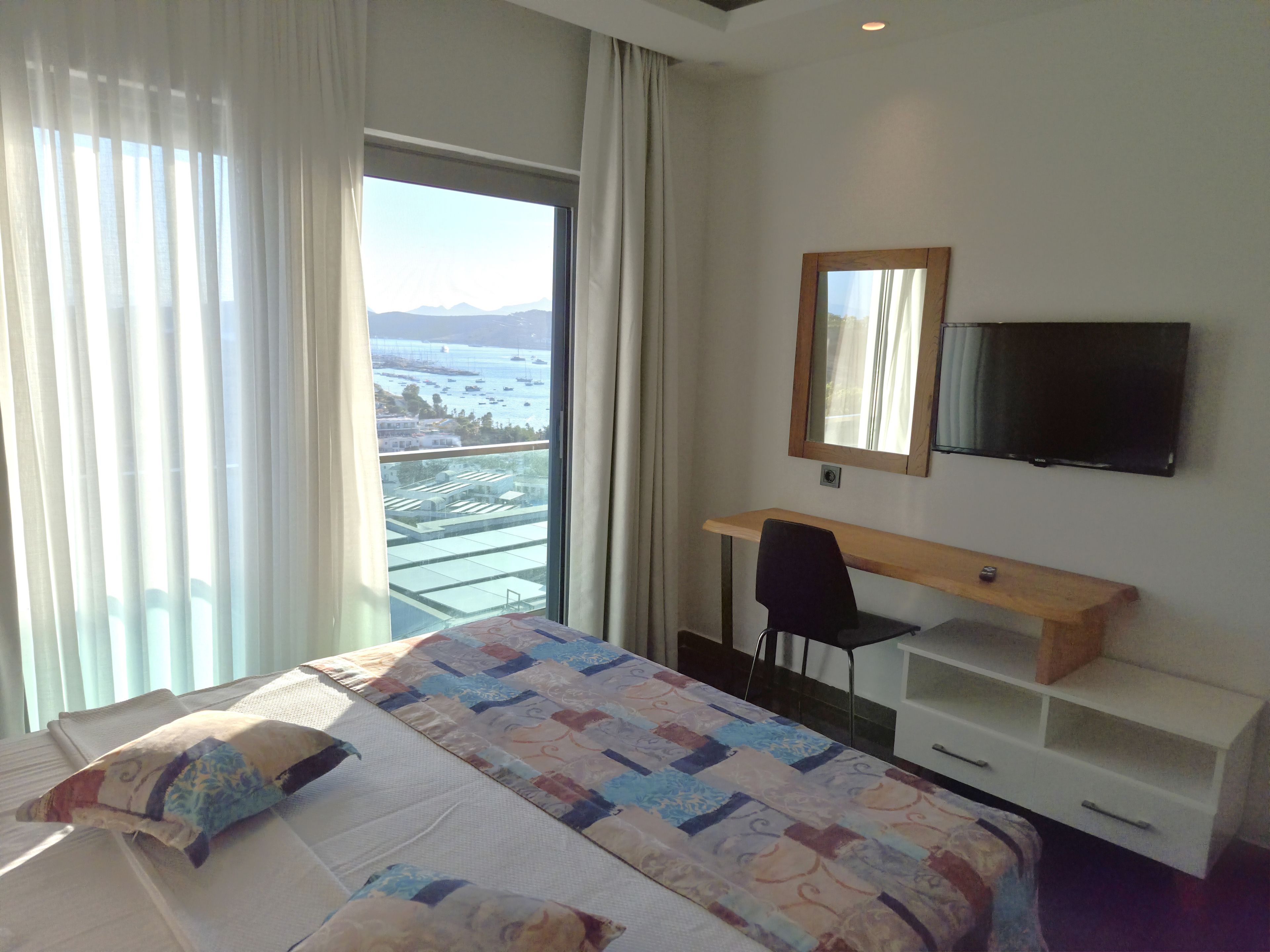 Quarto de hotel com uma cama grande, uma escrivaninha, uma cadeira, uma televisão e vista para o mar e as montanhas.