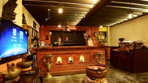 Uma área de recepção com televisão exibindo um canal esportivo, um balcão de atendimento e decoração tradicional, incluindo uma grande tigela de latão e luminárias pendentes.