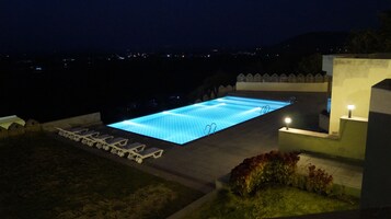 Piscina com espreguiçadeiras e área iluminada, cercada por um muro baixo e vegetação.