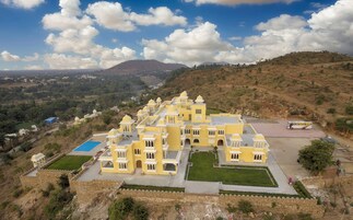 Um edifício grande e amarelo, com várias cúpulas e janelas em arco, rodeado por um gramado bem cuidado e uma parede de pedra.