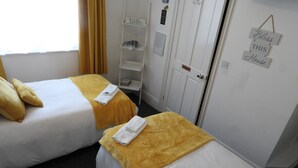 Quarto de hotel com duas camas, cada uma com um cobertor e almofadas amarelos, uma pequena estante com um livro, e uma porta com uma placa que diz 'Bless This House'.