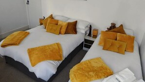 Quarto de hotel com duas camas, cada uma com roupa de cama branca e vários travesseiros amarelos. Há uma pintura na parede acima das camas, uma mesa de cabeceira com abajur e um urso de pelúcia na cama da direita.