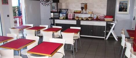 Um restaurante com cadeiras brancas e mesas com tampos vermelhos, um balcão com alimentos e um espelho decorativo fixado na parede.