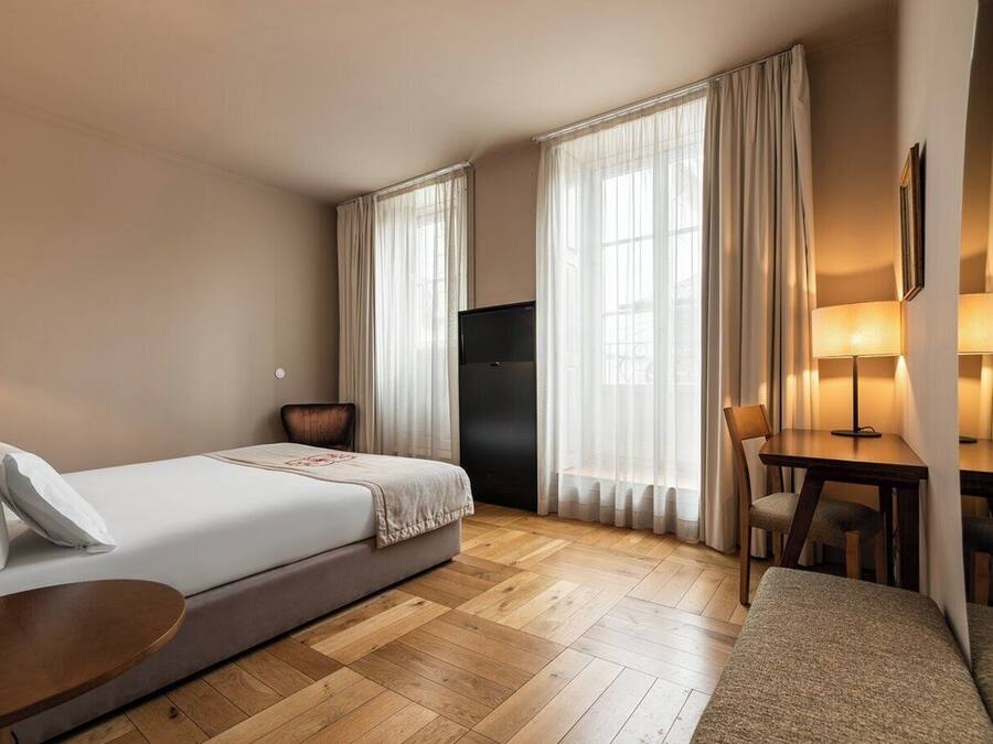 Quarto de hotel com uma cama, uma escrivaninha com cadeira, televisão e luminária.
