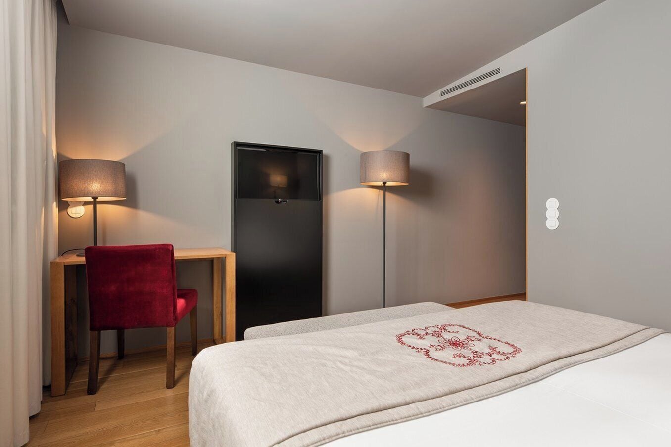 Quarto de hotel com cama, secretária, cadeira, duas luminárias e porta.