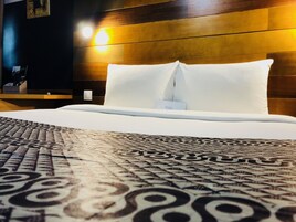 Cama bem arrumada com roupa de cama branca e colcha estampada, cabeceira de madeira e luminária fixada na parede.