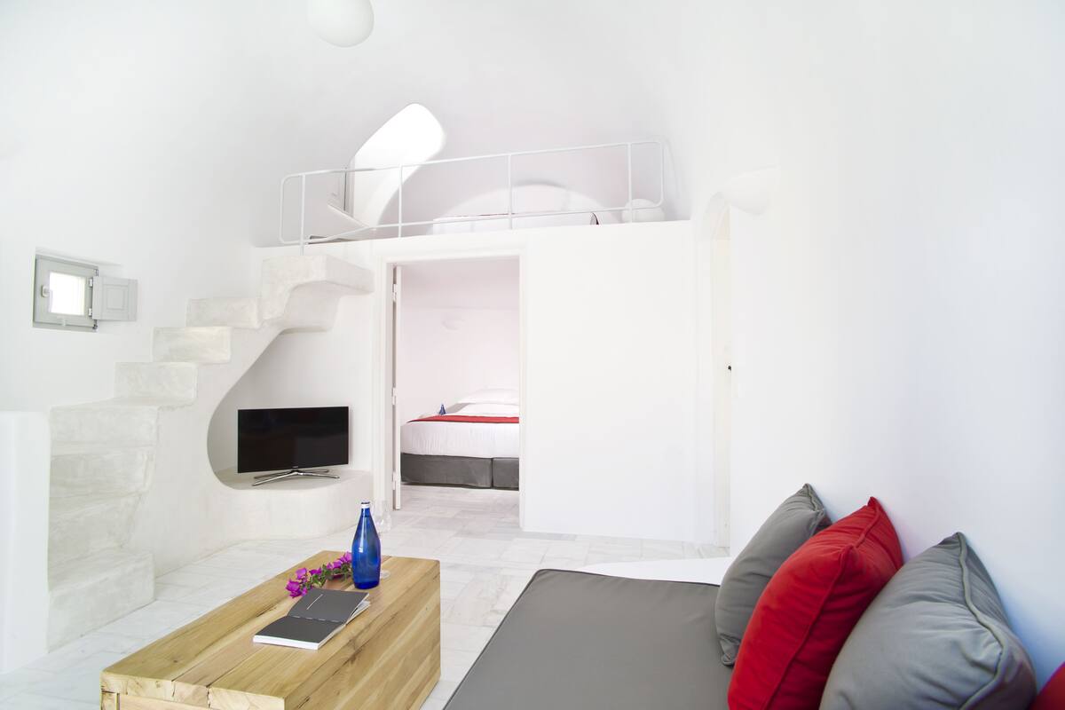 Quarto moderno com decoração em tons de branco, área de mezanino, televisão de tela plana, mesa de centro de madeira e cama com cobertor vermelho.