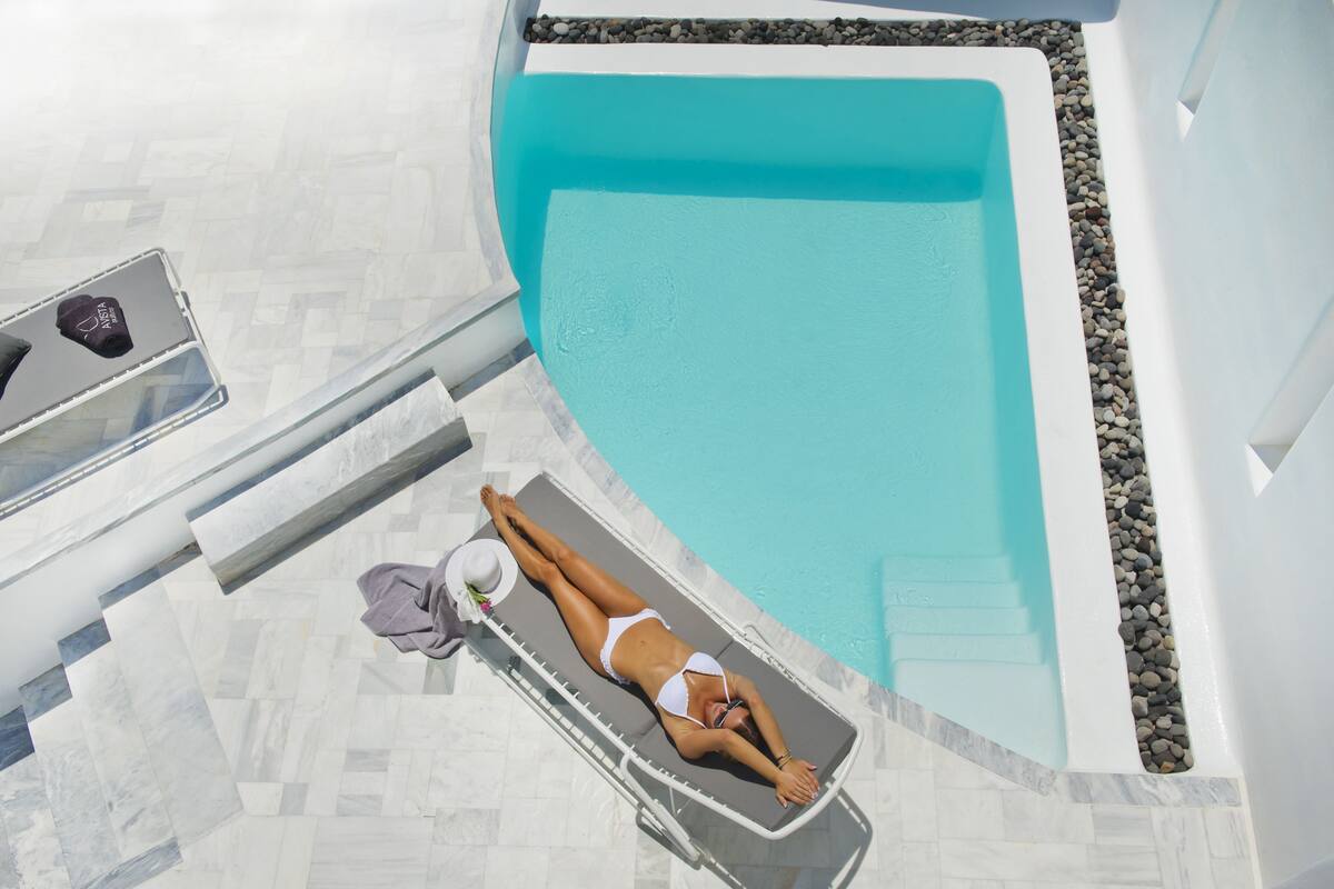 Mulher deitada em espreguiçadeira à beira de uma piscina, com piso de azulejos e parede branca.