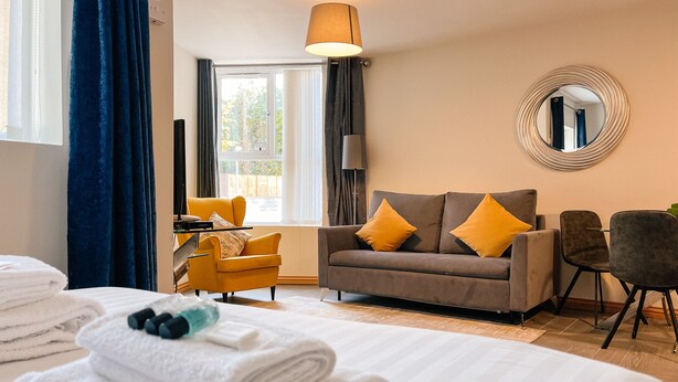 Quarto de hotel com cama, sofá, poltrona, luminária e janela com cortinas.