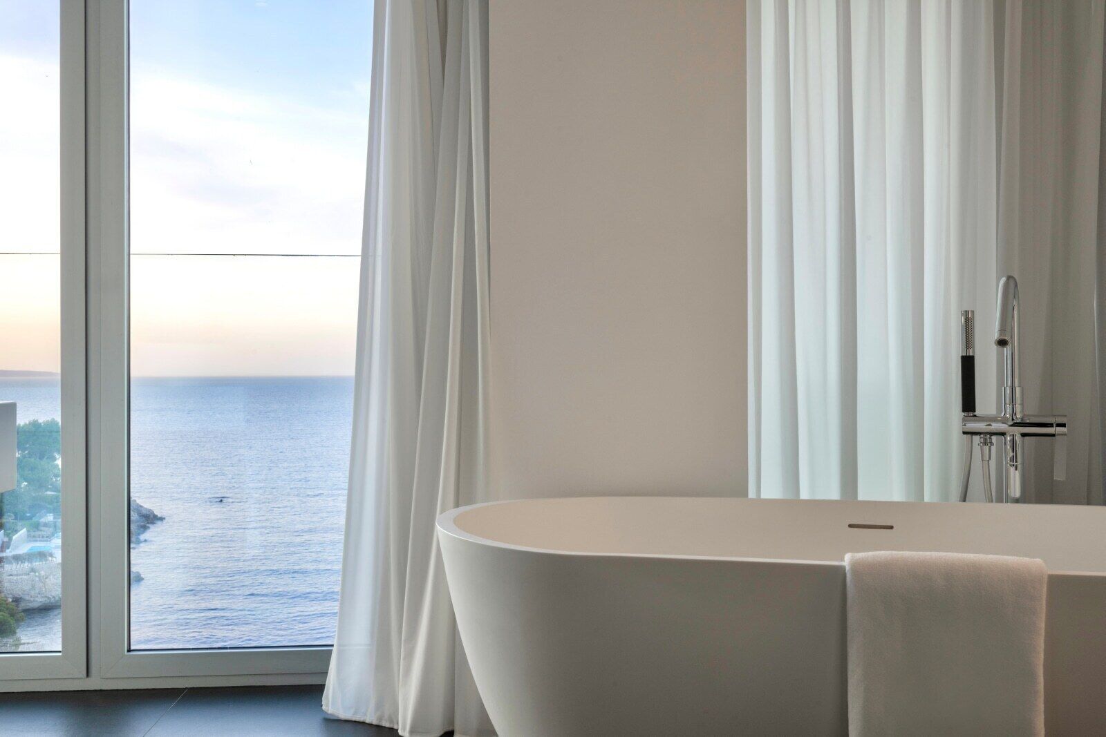 Um banheiro moderno com uma banheira de pés dourados, uma janela grande com cortinas e vista para o mar.