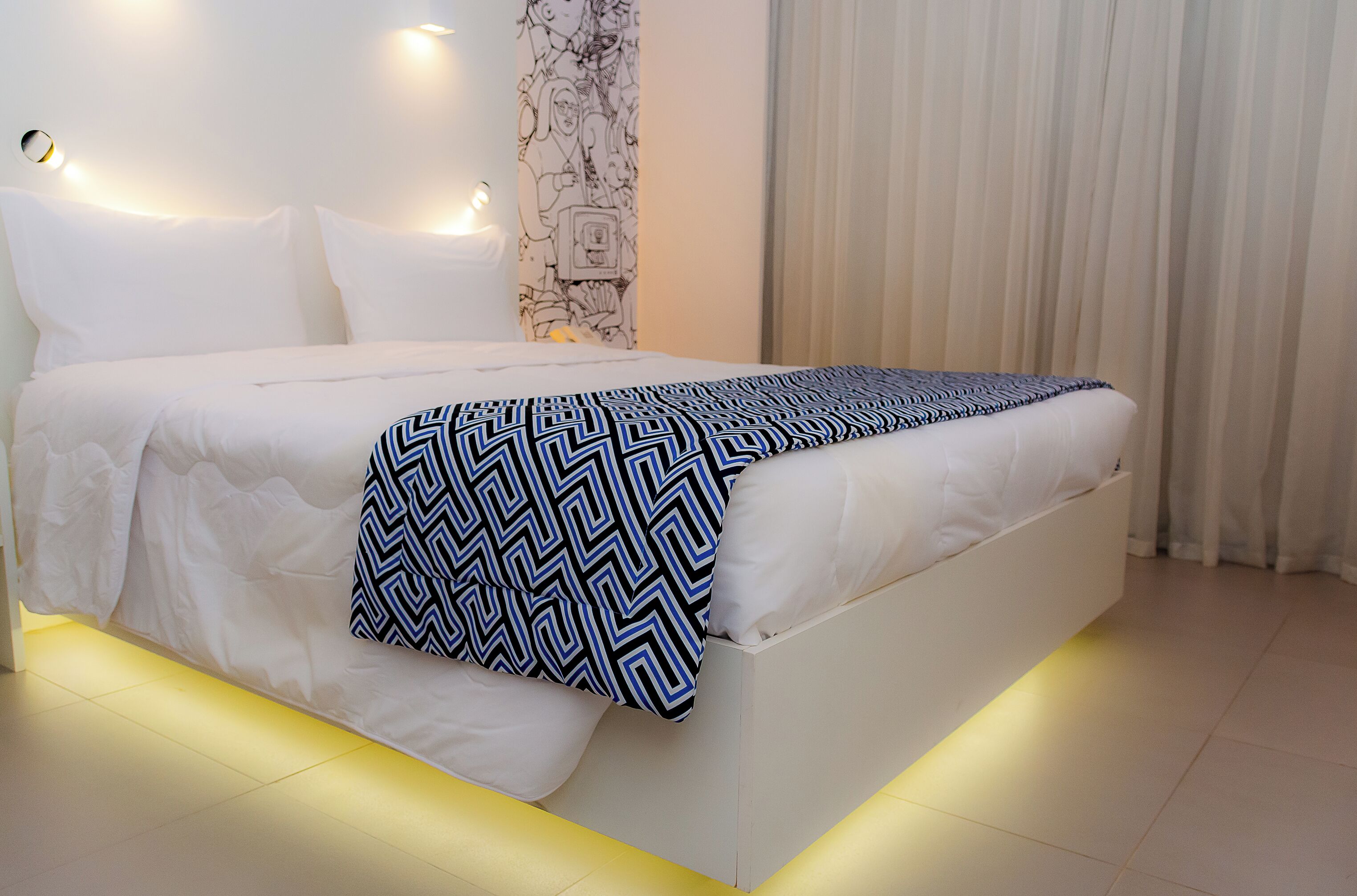 Quarto de hotel moderno com uma cama bem arrumada, manta com padrão e parede com um mural decorativo.