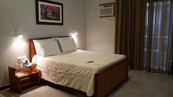 Quarto de hotel com cama, mesa de cabeceira, luminária, aparelho de ar condicionado e janela com cortinas.