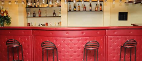 Uma área de bar com balcão revestido em vermelho, três banquetas pretas e prateleiras repletas de garrafas e copos.