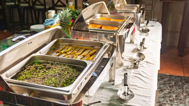 Uma mesa de buffet com diversos alimentos em recipientes de metal, uma abacaxi e uma toalha de mesa branca.