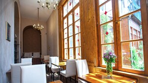 Uma área de refeições com mesas de madeira e cadeiras brancas, grandes janelas com vidros decorativos e um lustre.