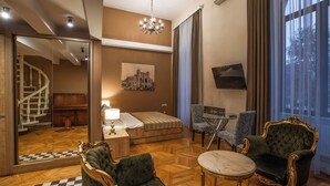 Quarto de hotel com cama, escrivaninha com luminária, cadeira, mesa redonda, televisão, espelho grande e escada.