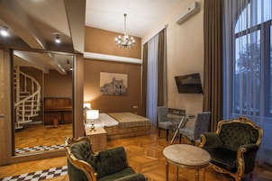 Quarto de hotel com cama, escrivaninha com luminária, cadeira, mesa redonda, televisão, espelho grande e escada.